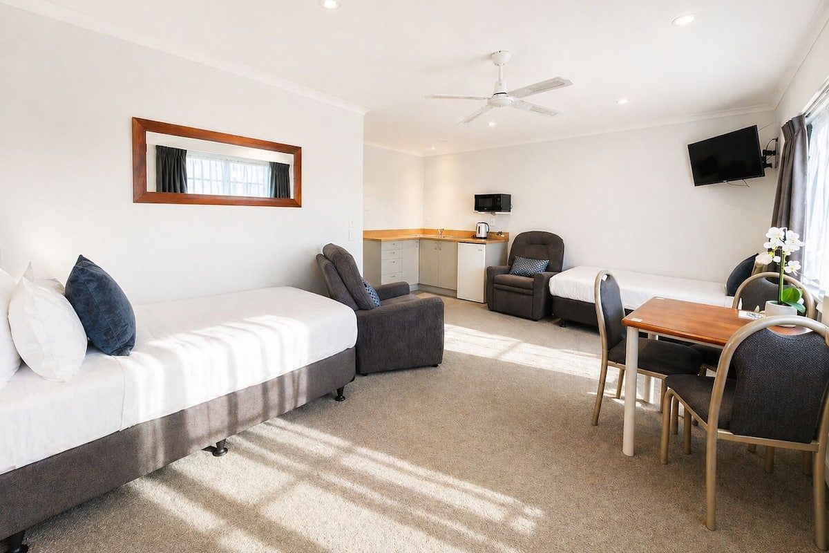 Quarto de hotel com cama, duas poltronas, uma mesinha, televisão, ventoinha no teto e uma cozinha compacta.