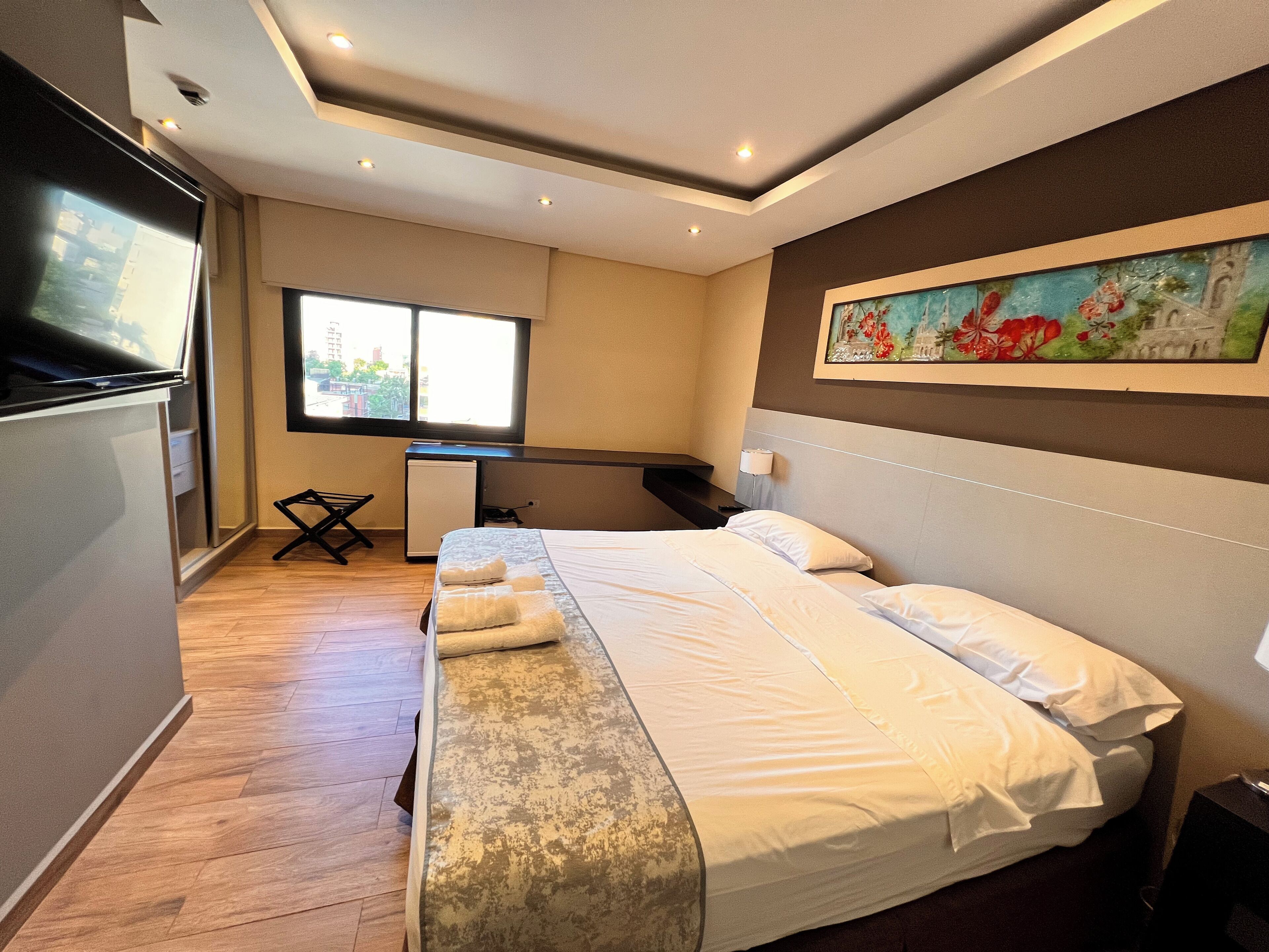 Quarto de hotel com cama, escrivaninha, televisão, janela e um quadro na parede.