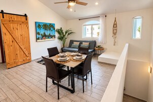 Uma área de refeições com mesa de madeira posta para quatro pessoas, sofá, planta em vaso e pintura de palmeiras.
