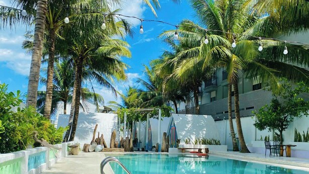 Piscina com água azul cristalina, rodeada por palmeiras e um edifício branco ao fundo.