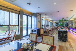 Uma ampla sala de jantar de hotel com tetos altos, janelas grandes e uma área de bar moderna.