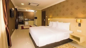 Quarto de hotel com uma cama grande, mesas de cabeceira, uma escrivaninha e uma cozinha compacta ao fundo.