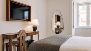 Quarto de hotel com cama, escrivaninha, cadeira, televisão, espelho e janela.