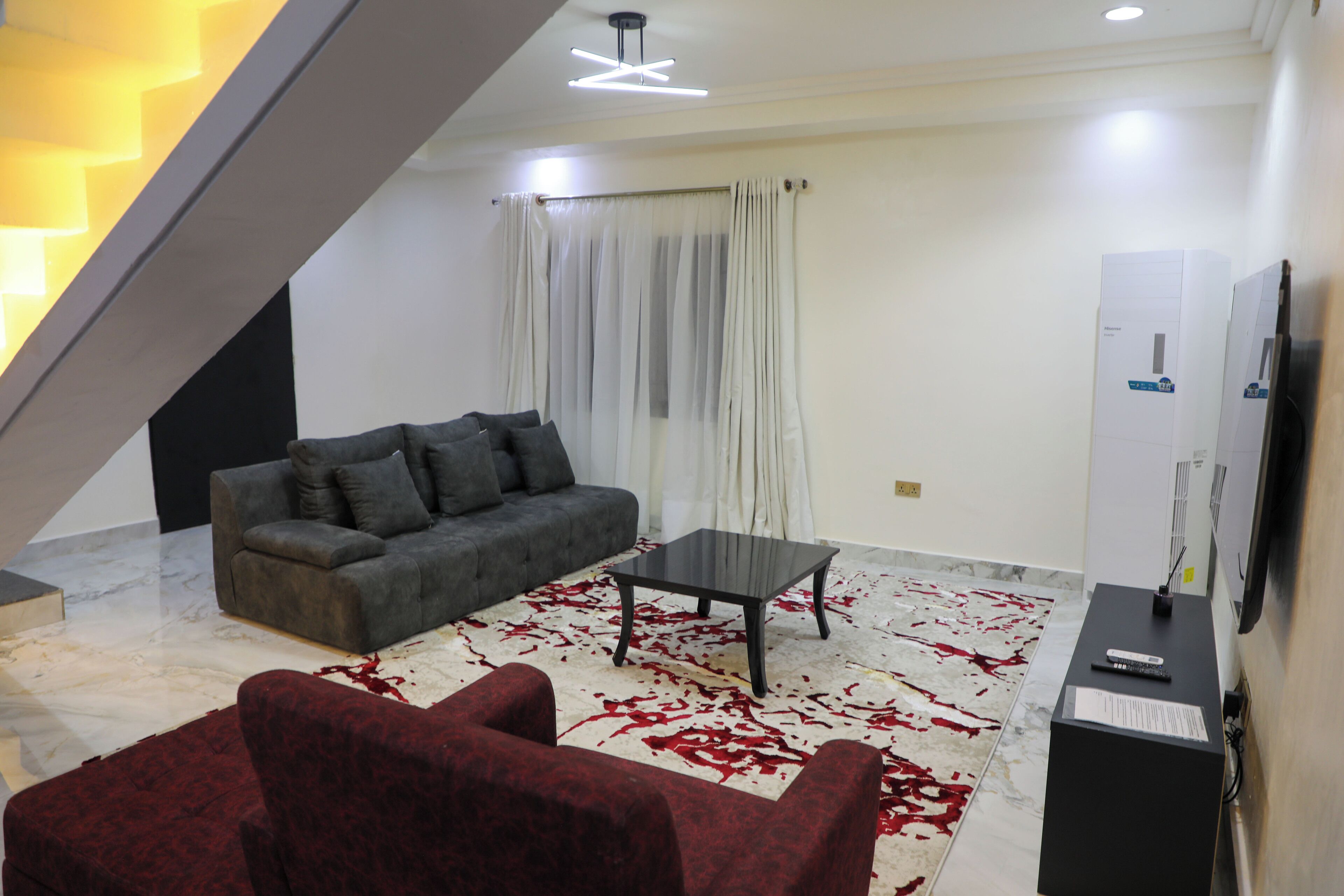 Uma sala de estar moderna com um sofá cinzento, poltronas vermelhas, uma mesa de centro preta e um tapete com padrão em branco e vermelho.