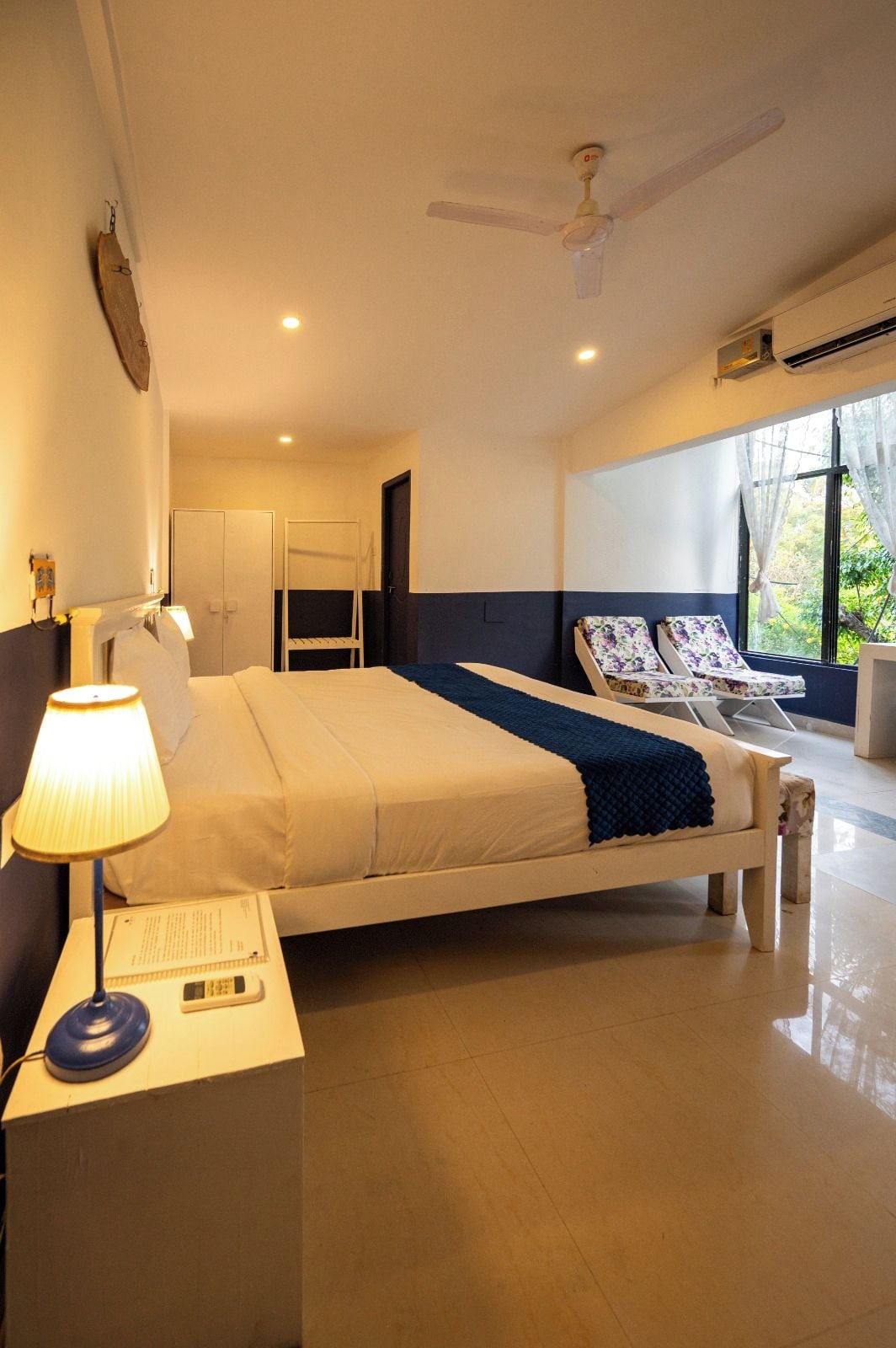Quarto de hotel com cama, luminária, mesa de cabeceira com controle remoto, ventilador de teto, janela com cortinas e vista para o verde lá fora.