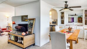 Uma sala de estar com sofá, mesa de centro com livros, área de jantar com mesa e cadeiras, e cozinha com eletrodomésticos.