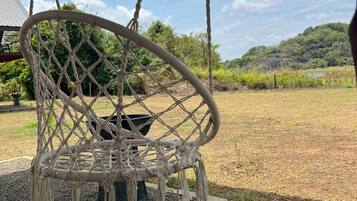 Uma cadeira balanço de vime branca suspensa por cordas, sobre um leito de cascalho, com céu claro e vegetação ao fundo.
