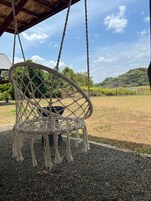 Uma cadeira balanço de vime branca suspensa por cordas, sobre um leito de cascalho, com céu claro e vegetação ao fundo.