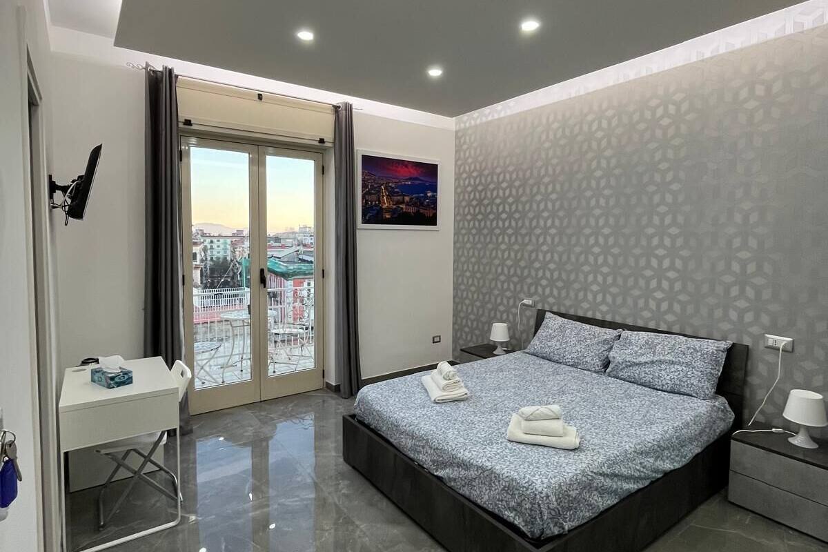 Quarto moderno com uma cama grande, uma escrivaninha e vista da cidade através de uma porta de vidro.