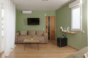 Uma sala de estar com sofá, mesa de centro, televisão, porta, uma mesinha com objetos decorativos e janela.