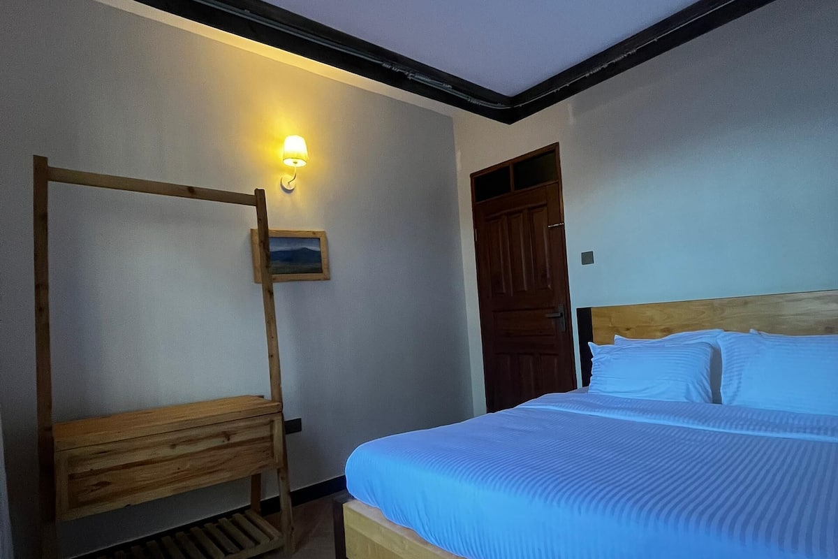 Quarto com estrutura de cama de madeira, um banco, luminária na parede e uma porta.