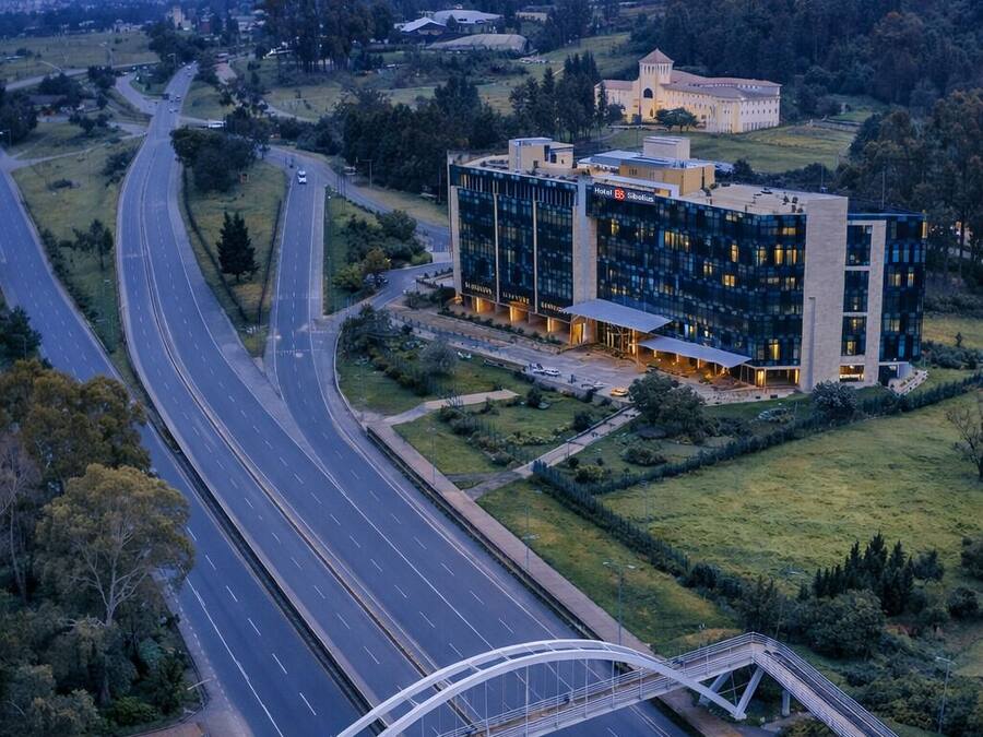 Hotel moderno com fachada de vidro, situado perto de uma rodovia e rodeado por vegetação.
