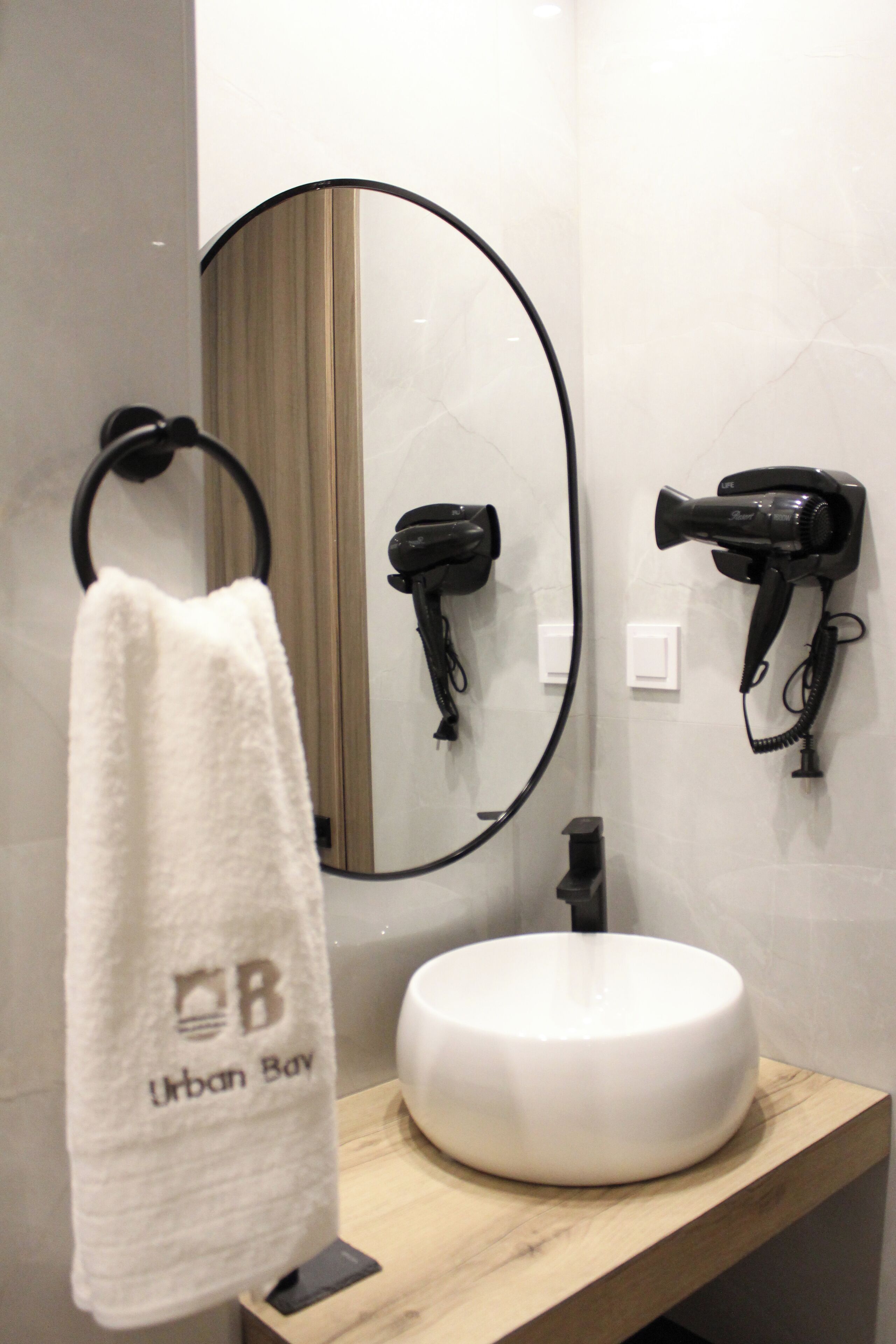 Uma casa de banho com uma pia branca, uma toalha pendurada num suporte e dois secadores de cabelo fixados na parede.