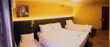 Quarto de hotel com duas camas, uma mesa de cabeceira com abajur, um quadro na parede e uma porta.