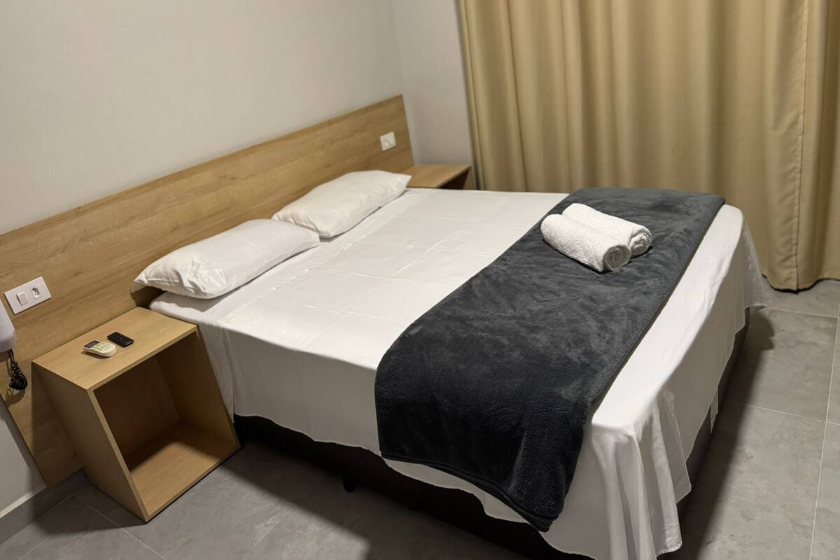 Cama bem arrumada com lençóis branco e cinza escuro, cabeceira de madeira, mesa de cabeceira com um telefone e uma cortina bege ao fundo.
