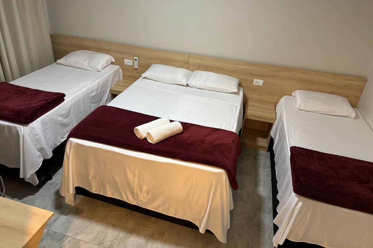 Quarto de hotel com três camas de solteiro, cada uma com roupa de cama branca e manta vermelha.