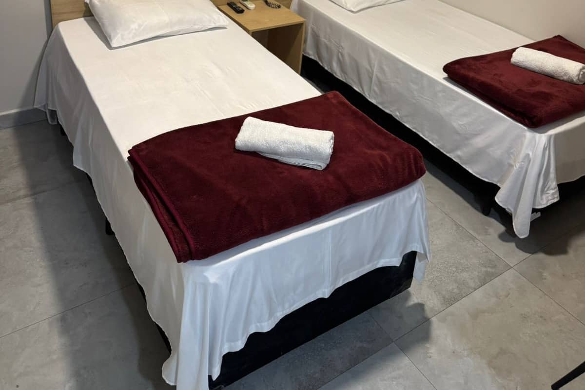 Quarto de hotel com duas camas de solteiro, cada uma com roupa de cama branca e manta vermelha. Há uma cabeceira de madeira, uma mesa de cabeceira com telefone e uma tomada na parede.