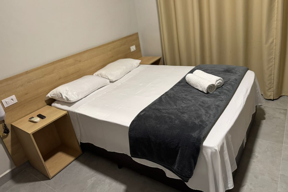 Cama bem arrumada com lençóis branco e cinza escuro, cabeceira de madeira, mesa de cabeceira com um telefone e uma cortina bege ao fundo.