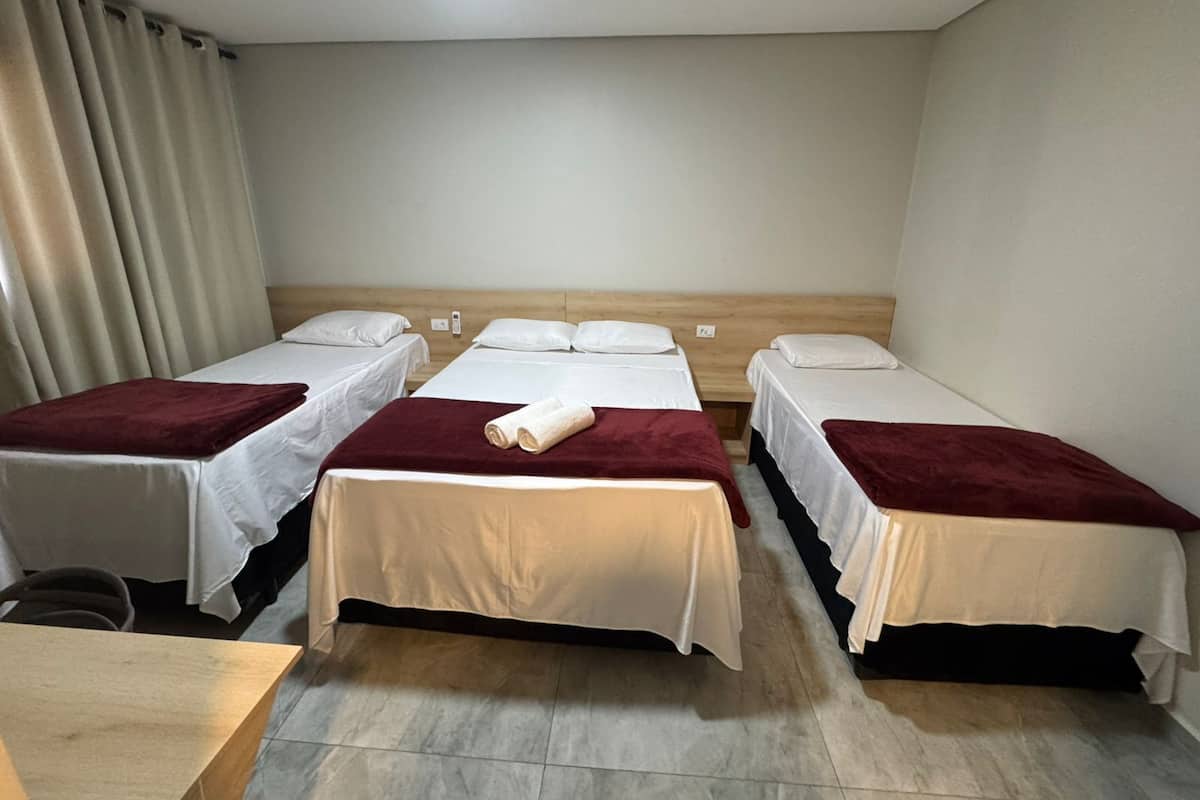 Quarto de hotel com três camas de solteiro, cada uma com lençol branco e manta vermelha.