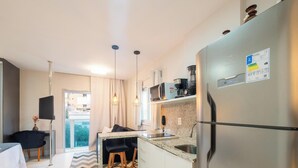 Cozinha compacta com geladeira, micro-ondas e cafeteira. Possui um pequeno espaço de refeições com mesa e cadeiras.