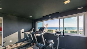 Uma academia com esteiras e bicicleta ergométrica, amplas janelas com vista para o oceano e um teto com iluminação embutida.