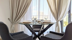 Uma sala de jantar com uma mesa de vidro redonda, duas cadeiras e vista para edifícios através de grandes janelas.