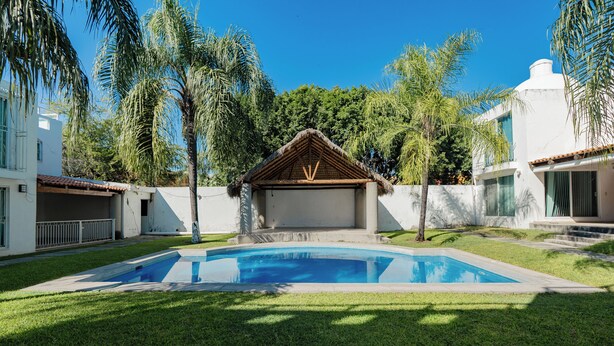 Piscina cercada por grama bem cuidada e palmeiras, com um edifício ao fundo apresentando uma estrutura de telhado de palha.