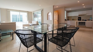 Uma sala de estar moderna com uma mesa de jantar de vidro e cadeiras pretas, uma cozinha com armários brancos e janelas grandes que oferecem uma vista.
