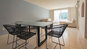 Uma sala de estar moderna com uma mesa de jantar de vidro e cadeiras de estrutura preta, um sofá branco e uma grande janela com vista para a cidade.