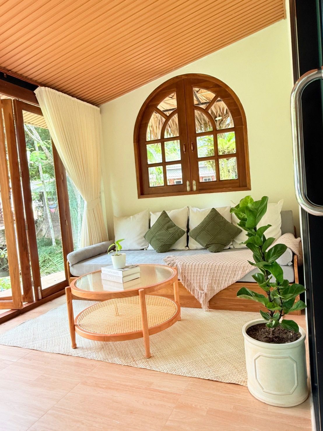 Um quarto com teto de madeira, uma grande janela em arco, um sofá com almofadas, uma mesa redonda com cadeiras e um vaso de plantas.