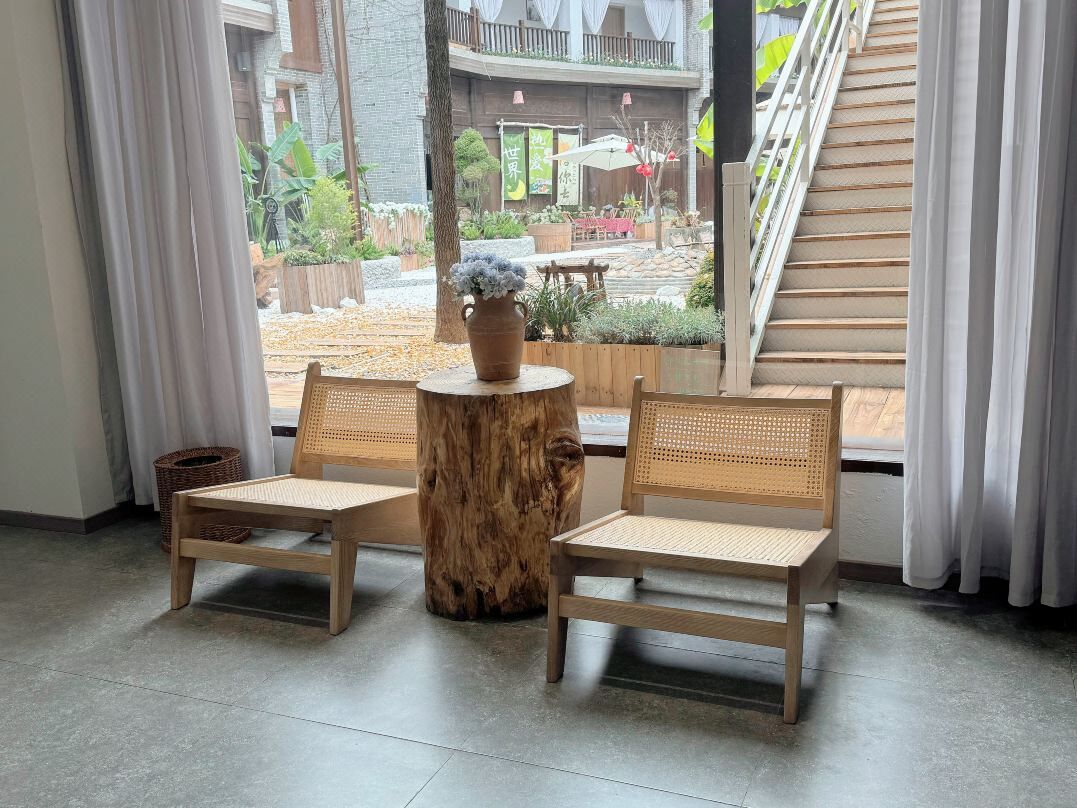 Quarto com duas cadeiras de madeira e uma mesa redonda de madeira, uma janela grande com cortinas e vista para um pátio externo com plantas e mobiliário de jardim.