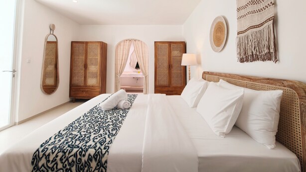 Quarto de hotel com uma cama grande, dois armários de madeira, um banheiro com pia e espelho, e um detalhe de macramê fixado na parede.