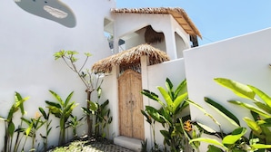 Um edifício branco com entrada de telhado de palha, rodeado por plantas tropicais e um chão de azulejos estampados.