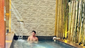 Um homem em uma banheira de hidromassagem com uma parede de pedra e plantas de bambu ao fundo.