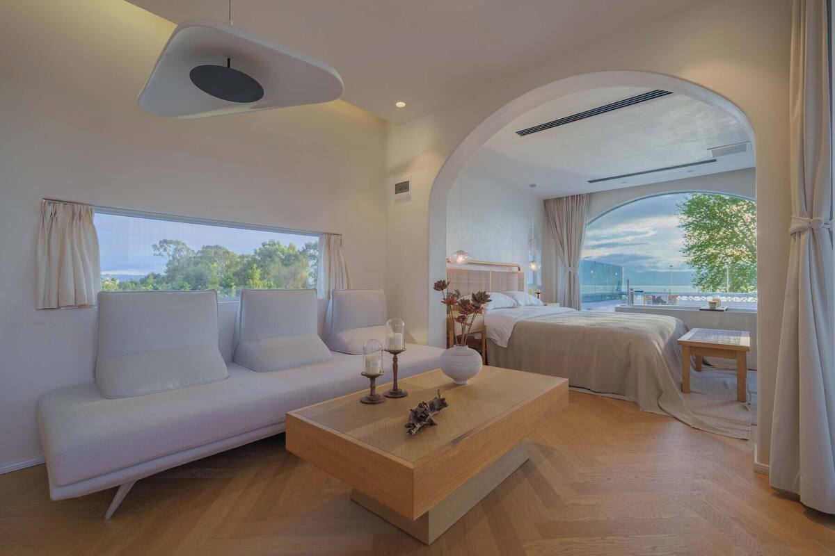 Quarto de hotel moderno com uma janela grande, um sofá, uma mesa de centro de madeira e uma cama.