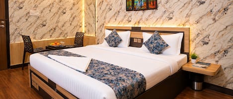 Quarto de hotel com uma cama grande, cabeceira de madeira, paredes com padrão marmorizado, um quadro sobre a cama, uma mesinha de cabeceira e uma cadeira.