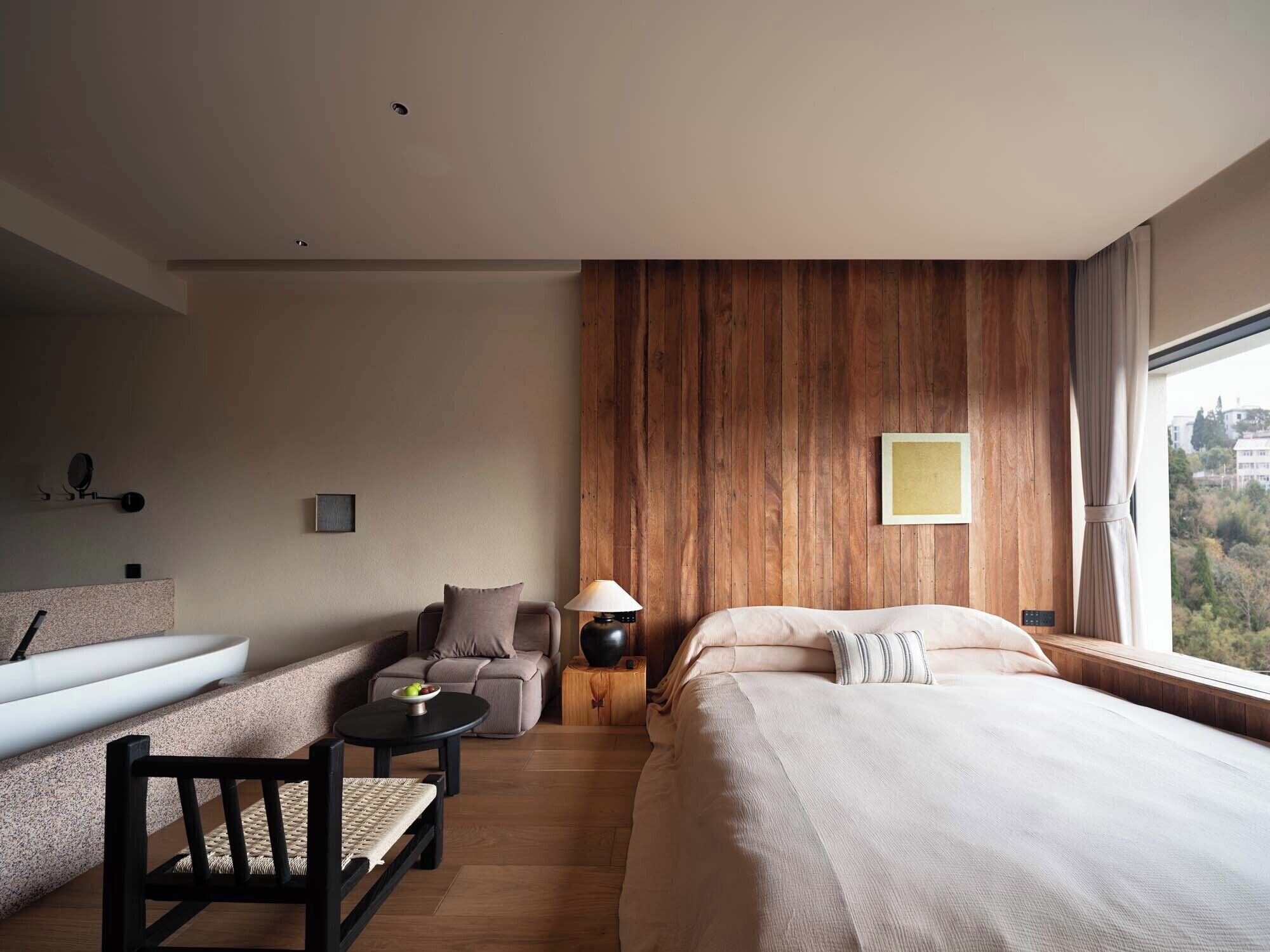 Quarto de hotel moderno com uma cama grande, um sofá, uma mesa pequena e vista para a cidade pela janela.