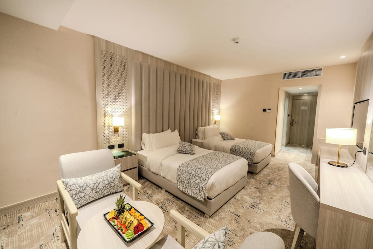 Quarto de hotel com uma cama grande, duas mesas de cabeceira, uma escrivaninha e uma cadeira. Há uma cesta de frutas na cama e uma luminária na escrivaninha.