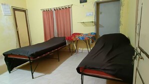 Quarto com duas camas, uma mesa pequena com toalha de mesa colorida, uma janela com cortinas rosa e uma porta.