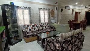 Uma sala de estar com sofá, mesa de centro de vidro, mesa de jantar com cadeiras, aparador com portas de vidro e um vaso de planta.