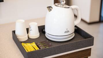 Uma chaleira de hotel branca com tampa sobre uma bandeja, acompanhada por duas chávenas brancas e um conjunto de saquetas de chá.