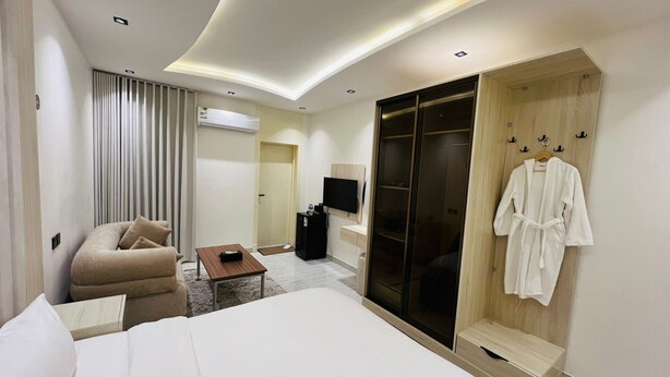 Quarto de hotel moderno com uma cama grande, área de estar com sofá e poltrona, televisão, guarda-roupa e roupão pendurado na parede.