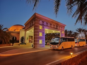 Entrada de hotel com galeria coberta, palmeiras e dois ônibus estacionados na frente.