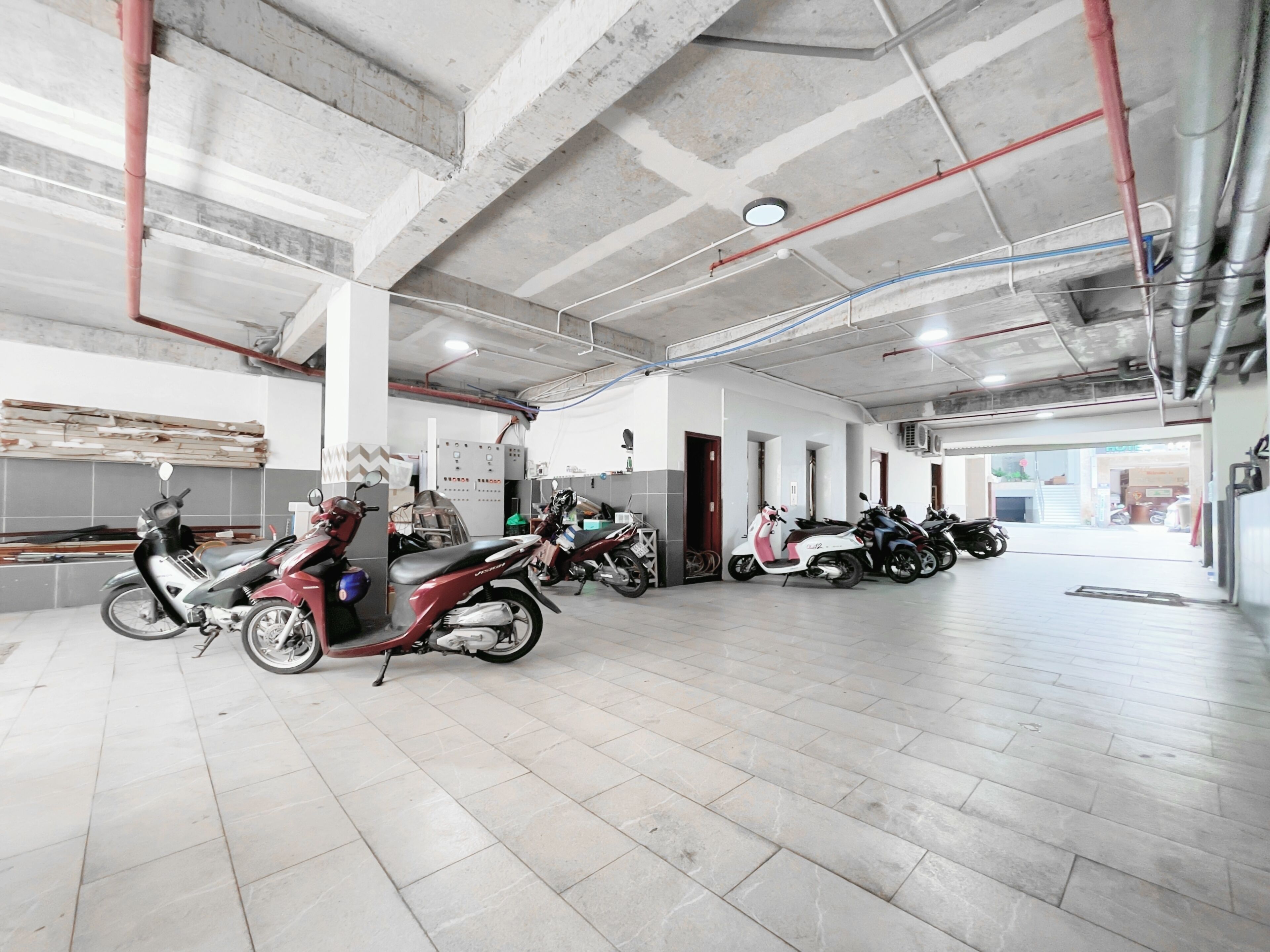 Um amplo estacionamento interno com várias scooters estacionadas, teto de concreto com vigas aparentes e piso de azulejos.