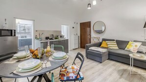 Uma sala de estar moderna com um sofá cinzento, uma mesa de jantar posta com pratos e copos, e uma cozinha ao fundo.