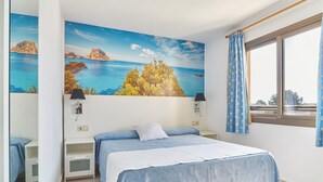 Quarto com cama, mesas de cabeceira, janela com cortinas e um mural de uma paisagem costeira.