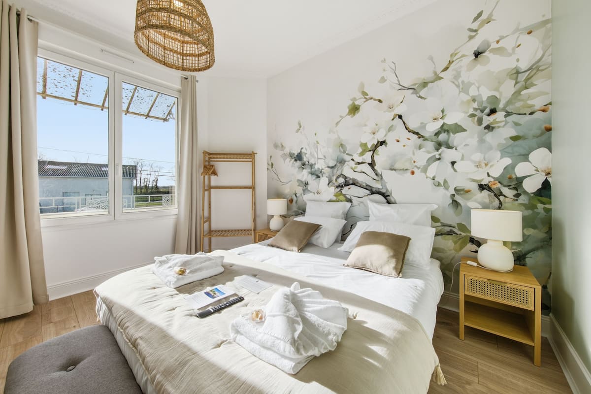 Quarto de hotel com uma cama grande, uma mesa de cabeceira com abajur, uma janela com cortinas e uma parede com um mural floral.