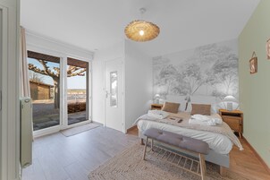 Quarto com cama, mesas de cabeceira, um banco, porta que dá para área externa e uma parede com mural de árvore.