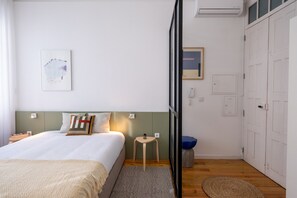 Quarto moderno com cama, mesa de cabeceira e um pequeno banquinho. É possível ver um guarda-roupa e uma porta ao fundo.