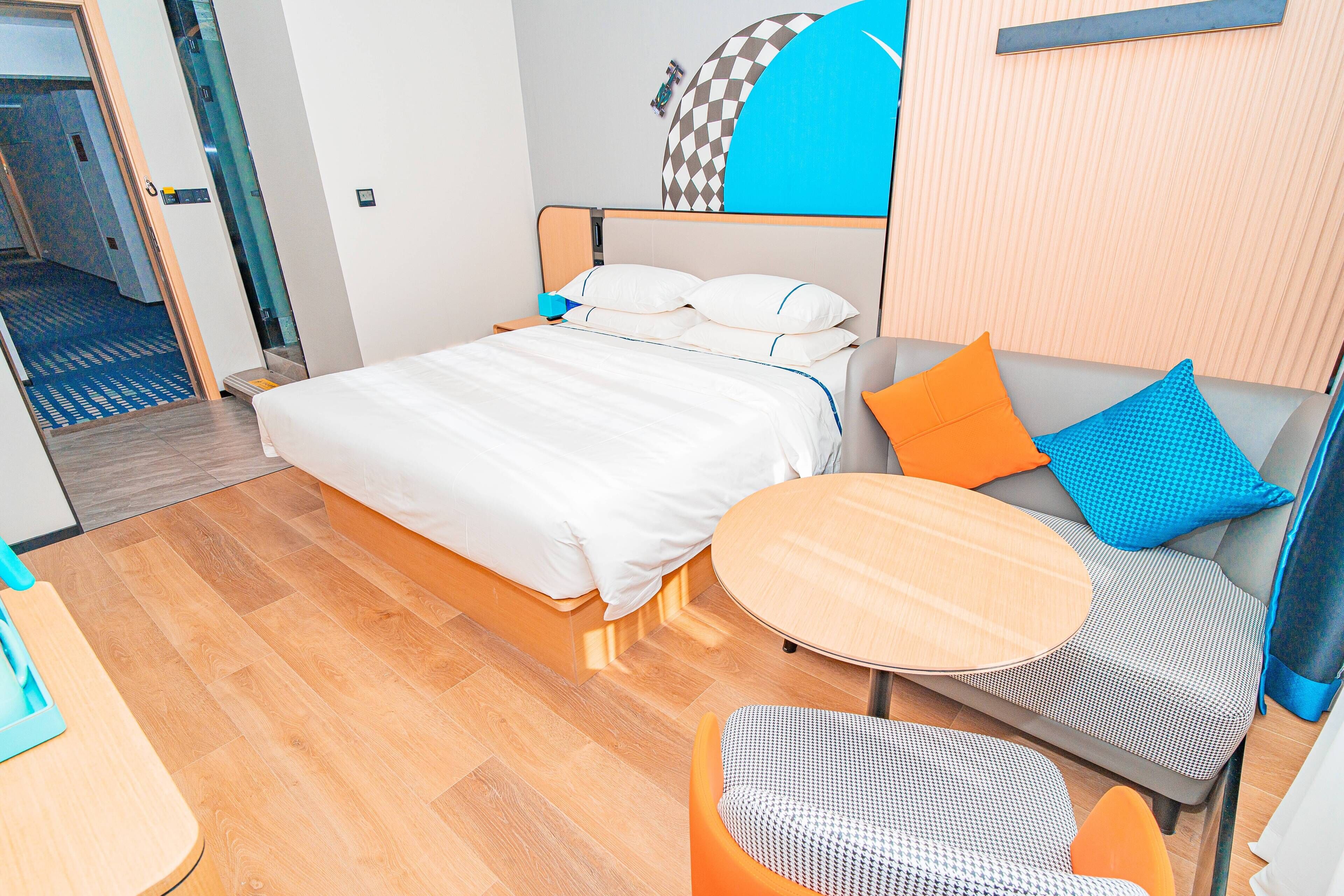 Quarto de hotel moderno com cama, mesa redonda, sofá com almofadas coloridas e decoração de parede xadrez.